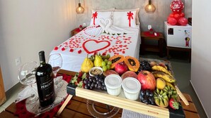 Quarto de hotel com cama decorada com um enfeite em formato de coração, bandeja de frutas e uma garrafa de vinho, e balões vermelhos pendurados no teto.