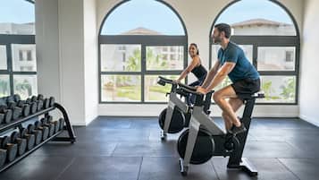 Duas pessoas pedalando em bicicletas ergométricas numa academia com grandes janelas e um suporte com halteres.