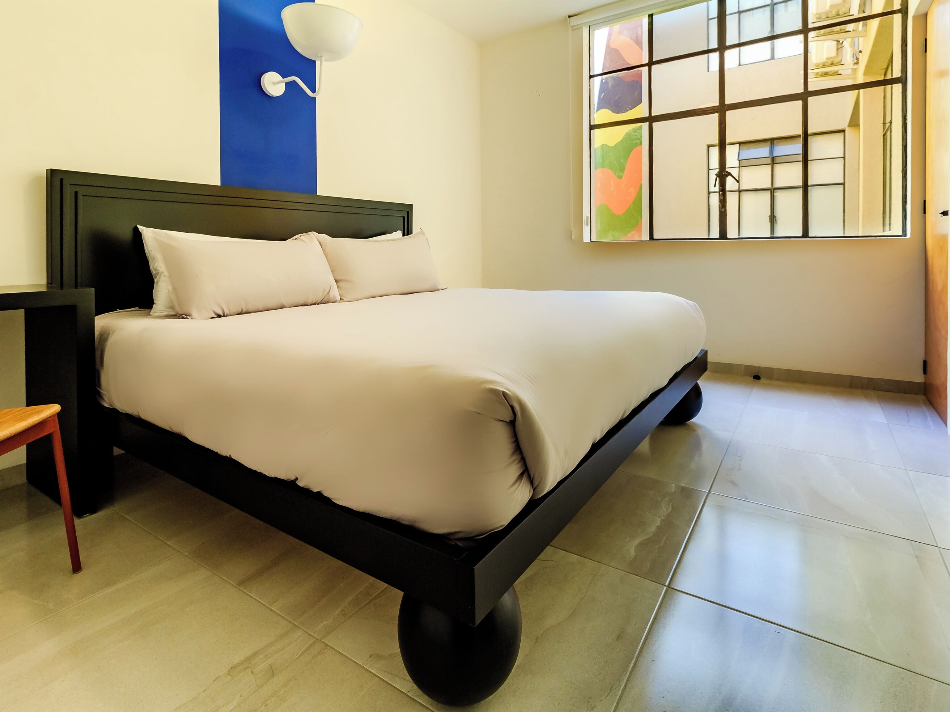 Uma cama bem arrumada, com cabeceira escura e roupa de cama branca, em um quarto com piso de azulejos e janela com vista colorida.