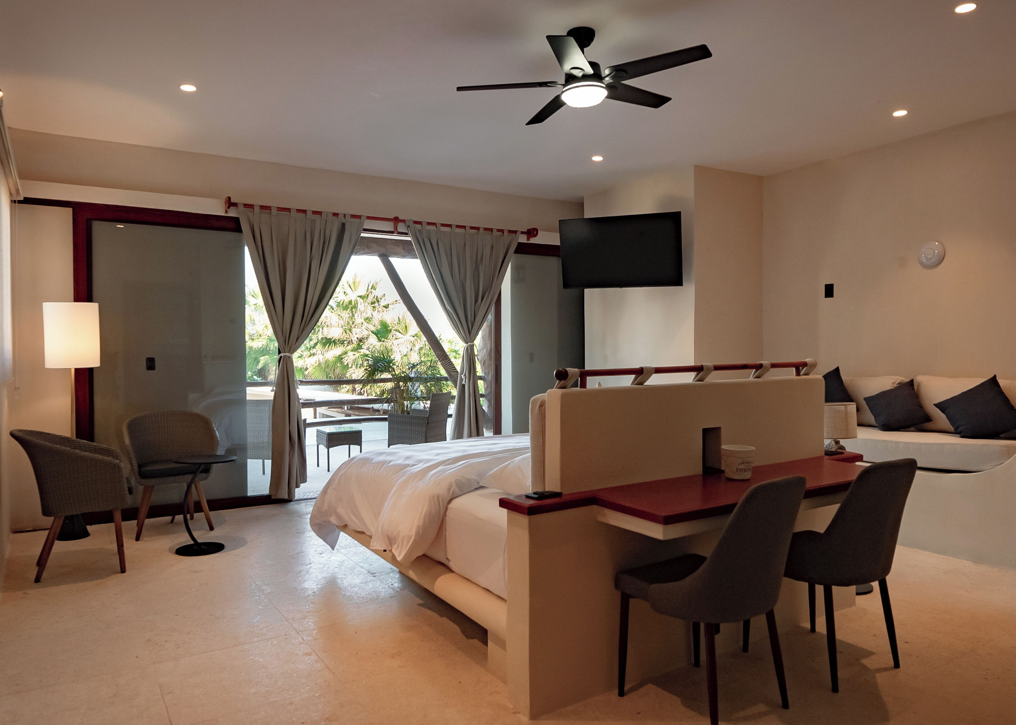 Quarto de hotel com cama, escrivaninha, cadeiras, televisão e varanda com cortinas.
