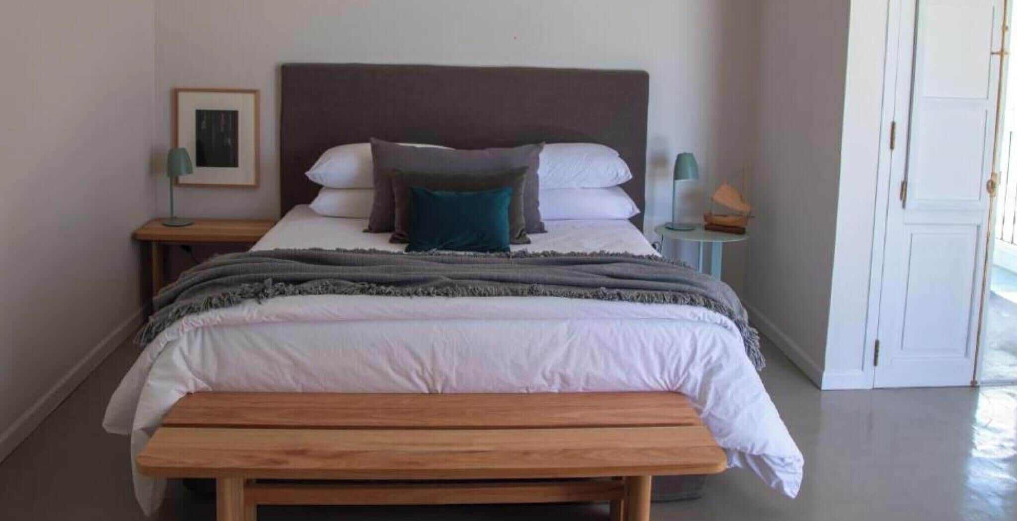 Uma cama bem arrumada com cabeceira, duas mesas de cabeceira com luminárias, um banco no pé da cama e um quadro pendurado na parede.