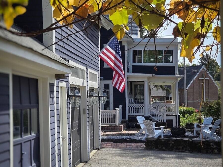 Uma casa de dois andares com varanda, uma bandeira americana e cadeiras Adirondack brancas.