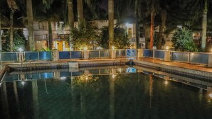 Piscina noturna com calçada revestida, cercada por palmeiras e edifícios.