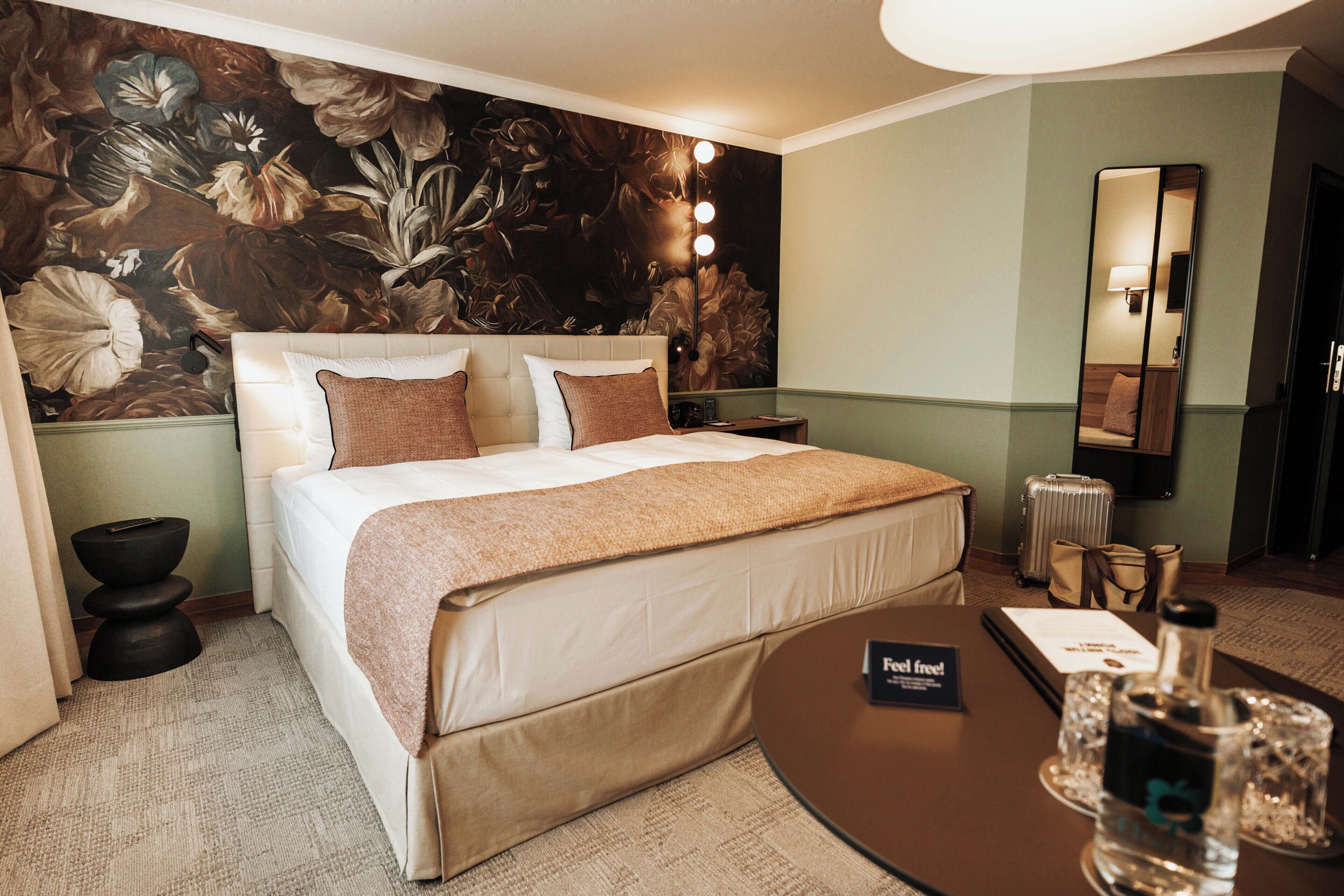Quarto de hotel com uma cama grande, uma mesa de centro redonda, um espelho fixado na parede e um mural floral.