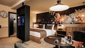 Quarto de hotel com cama, televisão, escrivaninha com abajur, cadeira e uma grande obra de arte floral na parede.