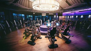 Uma academia com equipamentos de cardio, um painel central e amplas janelas.