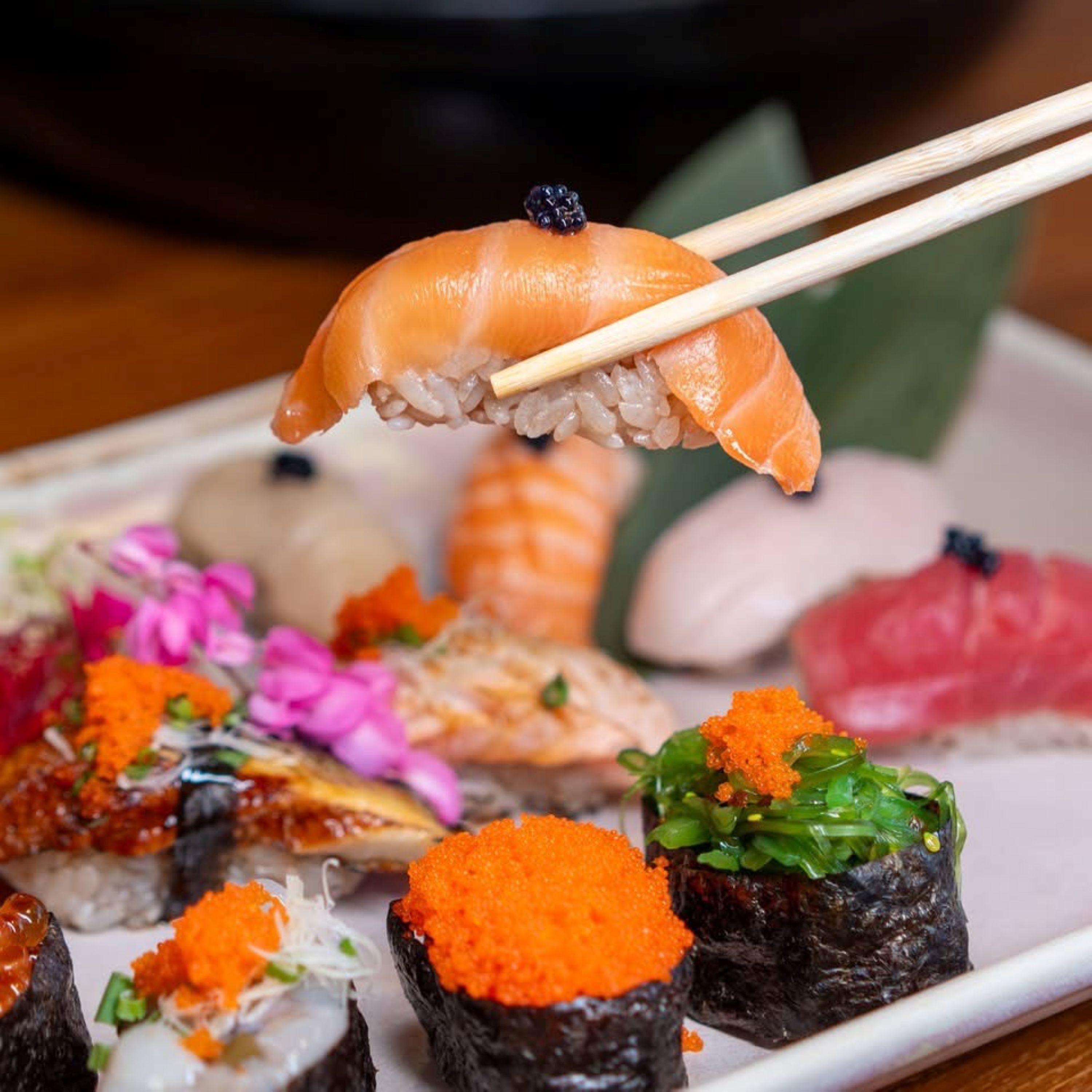 Diversos pratos de sushi em um prato, incluindo nigiri e enrolados maki, com hashis levantando um pedaço de nigiri.