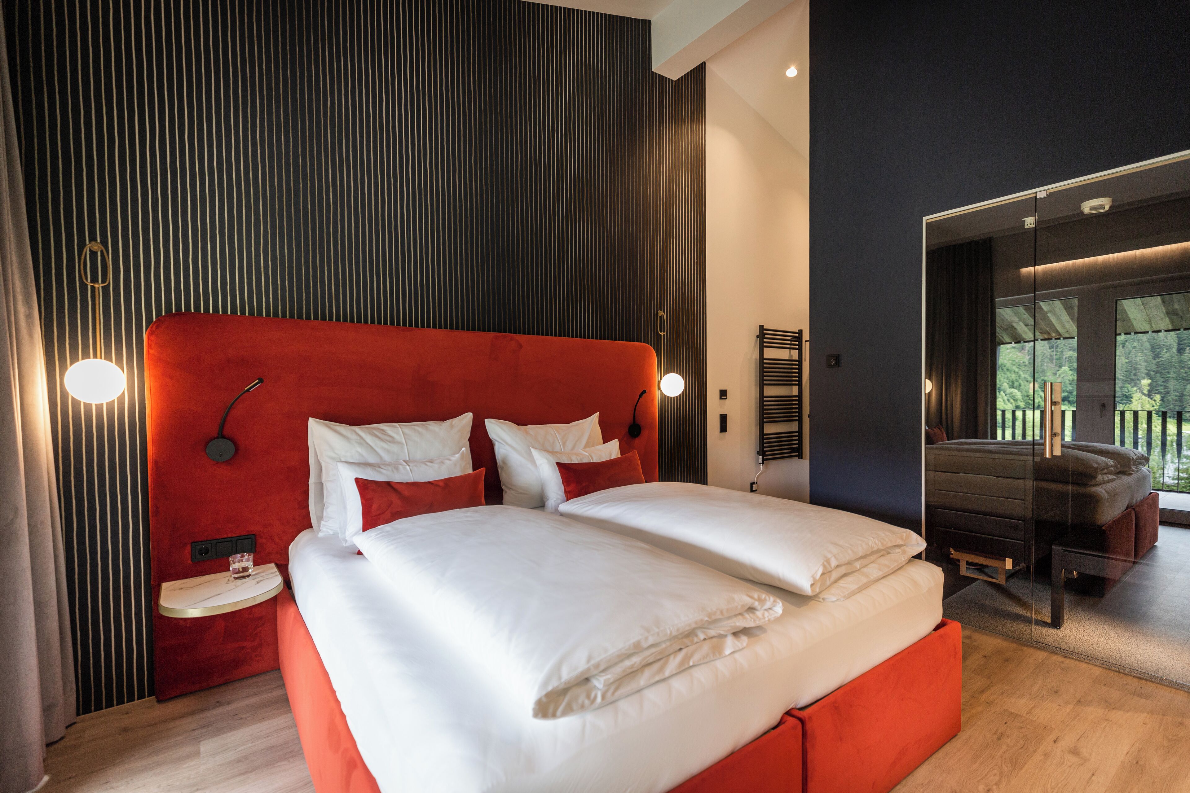 Quarto de hotel moderno com uma cama grande, uma mesinha de cabeceira, um espelho e vista para o exterior.
