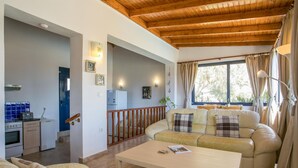Uma sala de estar aconchegante com um sofá bege, uma mesa de centro de madeira e uma área de cozinha visível ao fundo.