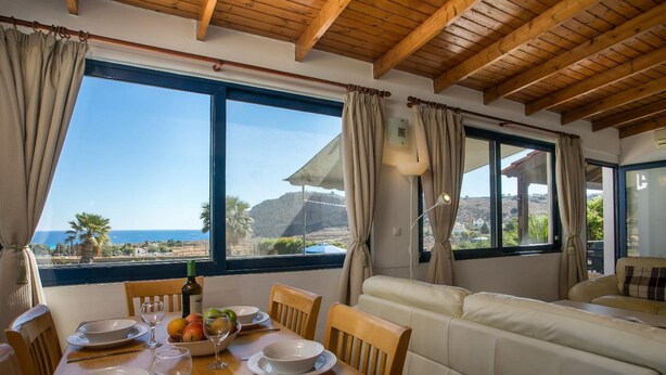 Uma área de jantar com uma mesa posta para dois, cadeiras de madeira, um sofá e janelas amplas com vista para o mar e colinas.