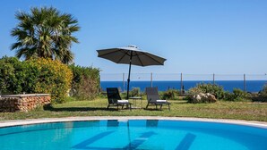 Piscina com espreguiçadeiras e guarda-sol, rodeada por vegetação e com vista para o oceano.