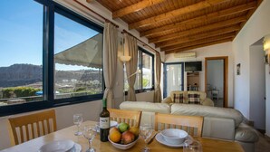Uma área de refeições com uma mesa posta para dois, uma tigela de frutas e uma garrafa de vinho. Há um sofá ao fundo e vista para a paisagem através de grandes janelas.