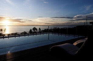 Piscina infinita com vista para o pôr do sol sobre o oceano, rodeada por palmeiras.