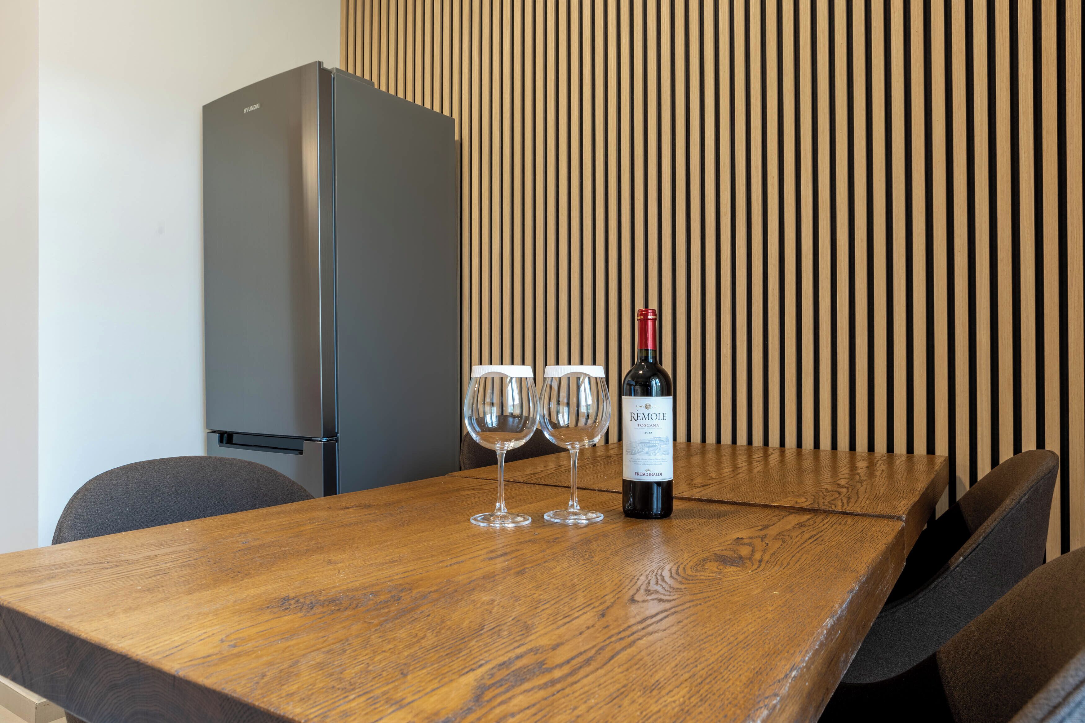Uma mesa de madeira com duas taças de vinho e uma garrafa, uma geladeira e um painel de parede de madeira.