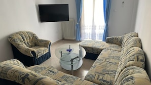 Uma sala de estar com sofá, mesa de centro de vidro e televisão de tela plana fixada na parede.