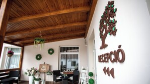 Uma área de recepção com teto de madeira, balcão de recepção, cadeiras pretas, parede branca com uma grande decoração de árvore e uma placa com a inscrição 'RECEPCIÓN'.