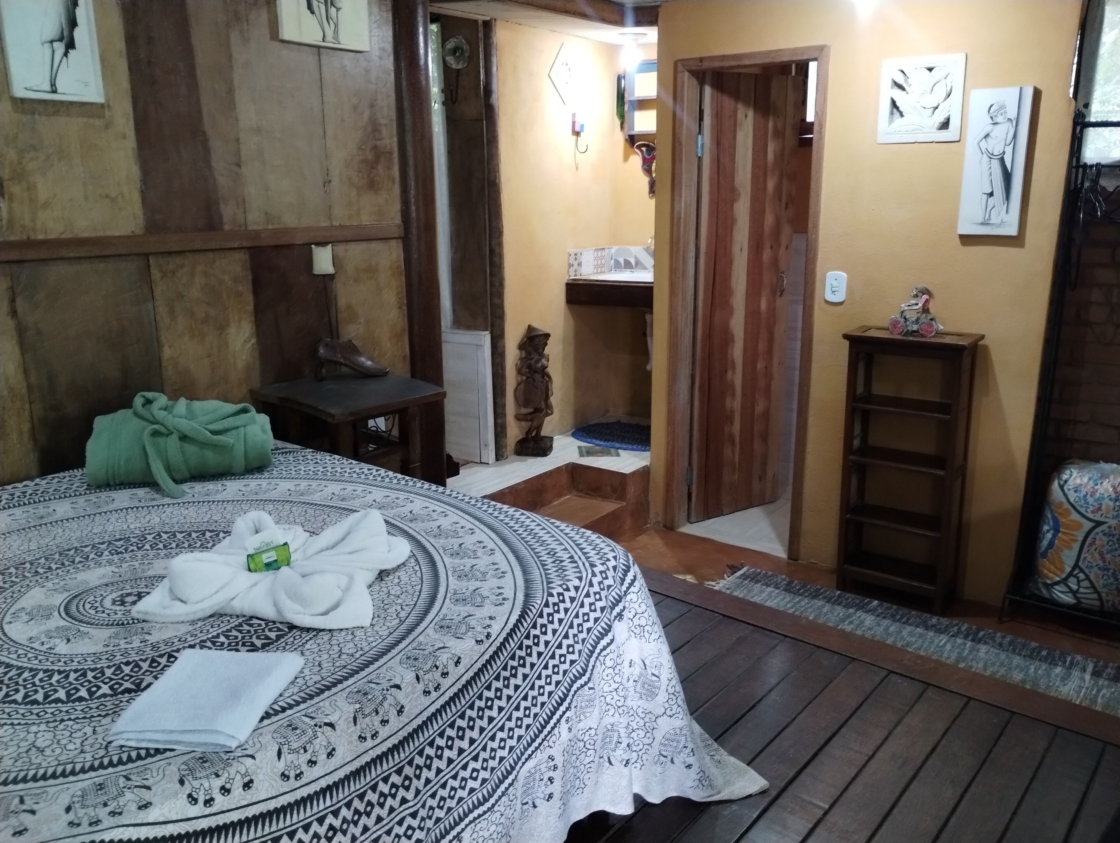 Quarto com cama coberta por uma colcha estampada, uma toalha verde e uma toalha branca dobradas na cama. Há uma porta de madeira que dá para outro cômodo, e um pequeno prateleira com uma estatueta em cima.
