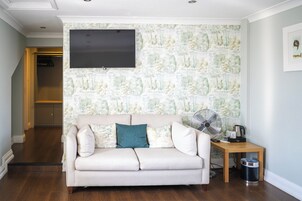 Uma sala de estar com um sofá bege, uma TV de tela plana na parede, um papel de parede estampado, uma mesinha de madeira com um ventilador e uma lixeira.