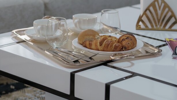 Uma bandeja de café da manhã com croissants e xícaras sobre uma mesa de centro branca em uma sala de estar moderna, com um sofá e arte decorativa na parede.