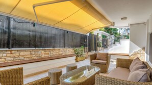 Área externa com assentos de vime, uma mesa de vidro e um toldo amarelo.