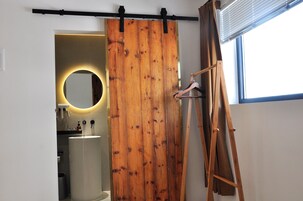 Um quarto com porta de madeira deslizante, espelho com luz circular, cavalete de madeira e janela com persianas.