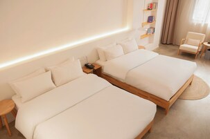 Quarto de hotel com duas camas, uma mesa de cabeceira com telefone, uma pequena mesa com abajur, uma cadeira e uma estante com livros.