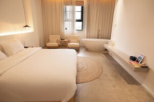 Quarto de hotel com uma cama grande, duas cadeiras, uma mesinha, uma banheira e uma prateleira com objetos.