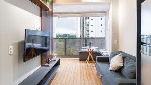 Sala de estar moderna com televisão de tela plana, sofá, mesa redonda pequena e varanda com vista para edifícios.