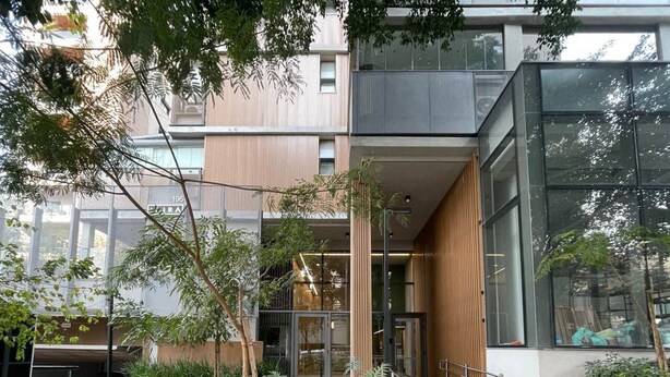 Um edifício moderno com fachada de vidro, entrada coberta e um pátio ajardinado com diversas plantas.