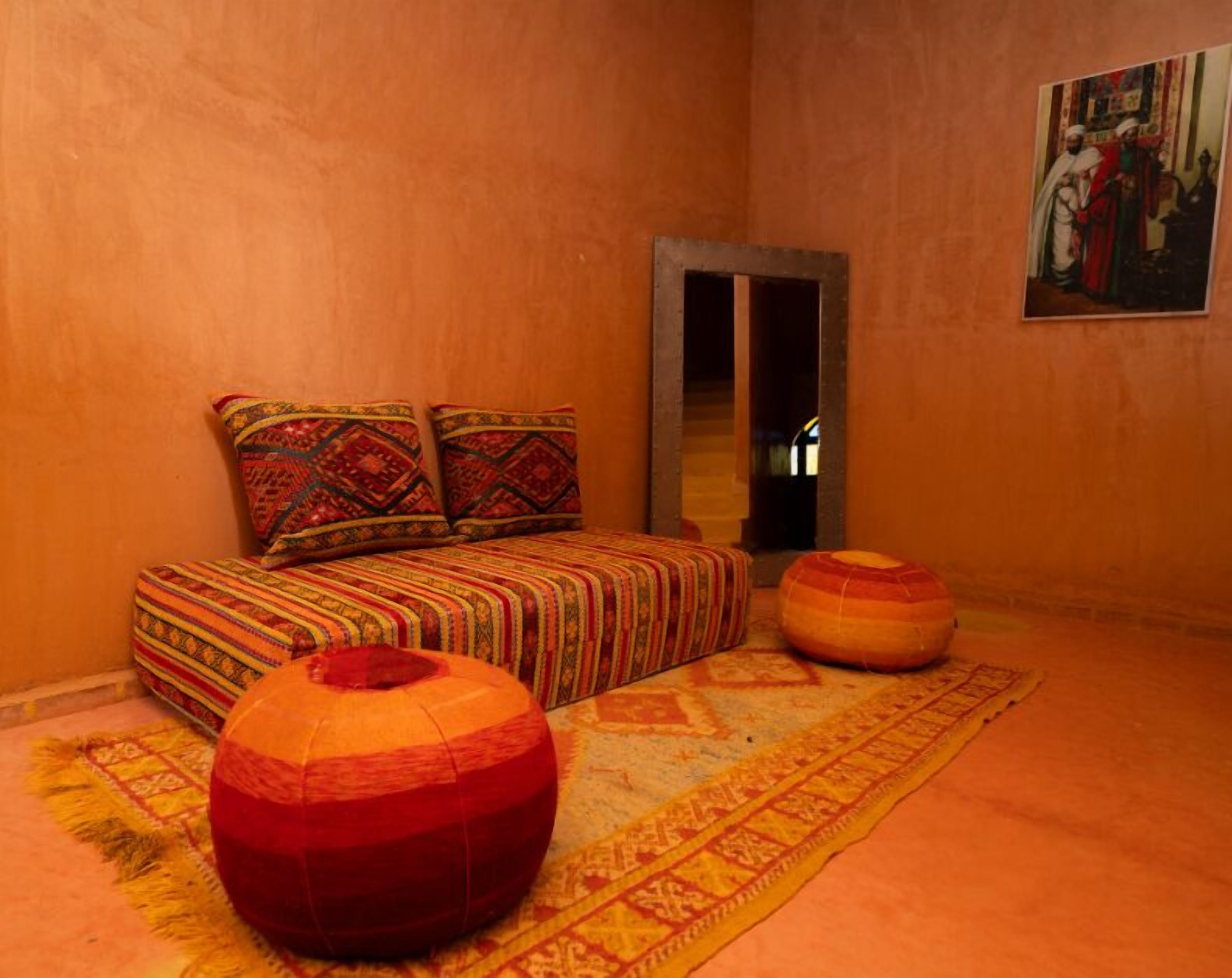 Quarto com cama, almofadas, tapete e um banquinho redondo.