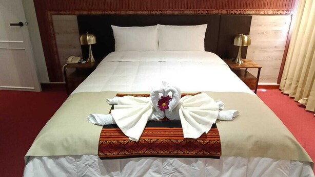 Quarto de hotel com uma cama grande, duas mesas de cabeceira com abajures, cabeceira de madeira e um tapete vermelho.