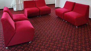 Quarto de hotel com carpete estampado em vermelho, três sofás vermelhos, uma mesa pequena com toalha branca e grandes janelas com vista para a cidade.