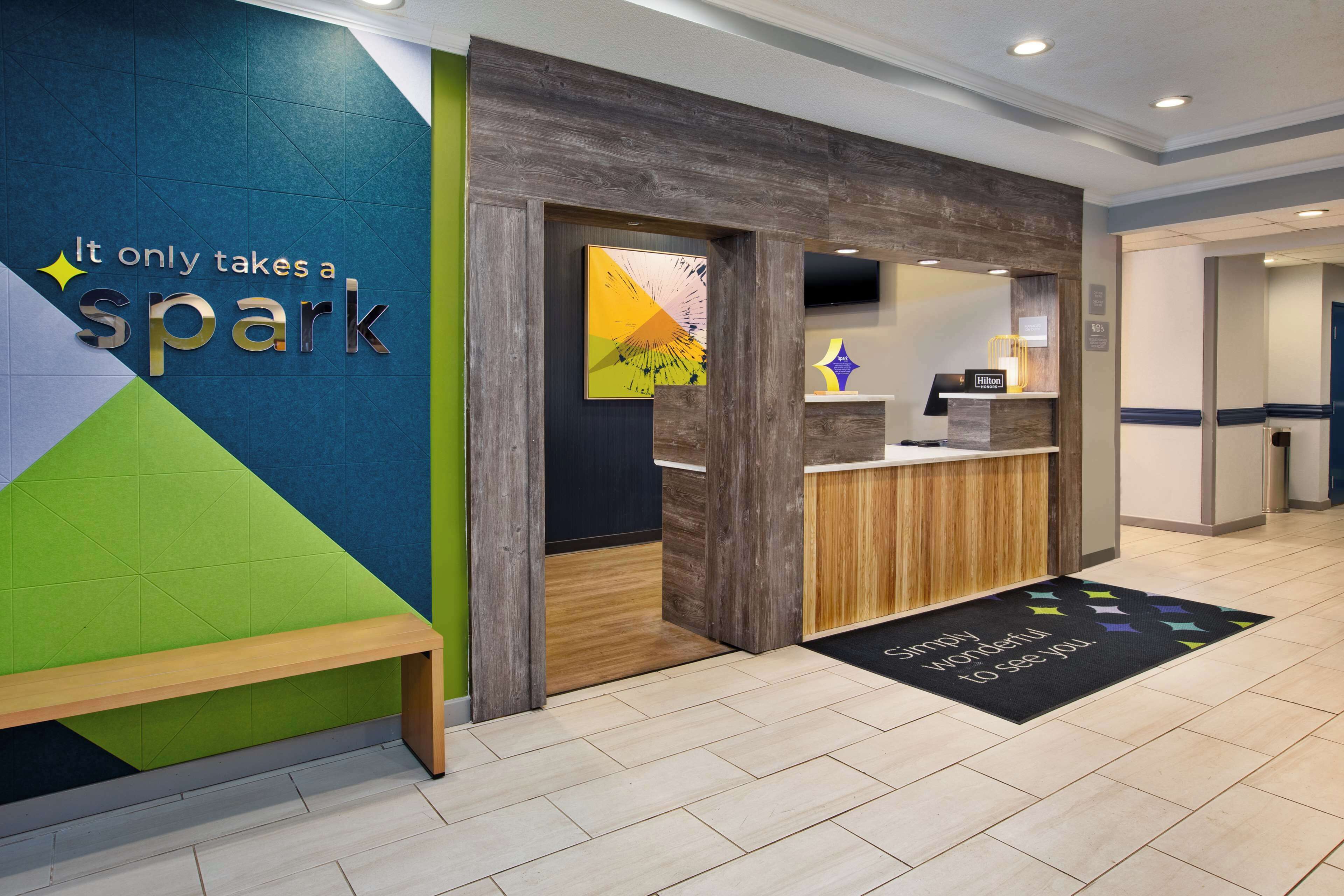 Uma área de recepção de hotel com design moderno, incluindo um balcão de madeira, uma parede colorida com a frase "It only takes a spark" e um banco.