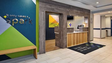 Uma área de recepção de hotel com design moderno, incluindo um balcão de madeira, uma parede colorida com a frase "It only takes a spark" e um banco.