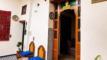 Quarto com piso xadrez, porta de madeira com uma placa, mesa pequena com cadeiras e luminária fixada na parede.