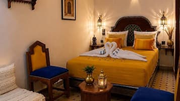 Quarto de hotel com cama, duas cadeiras, uma mesinha, abajur decorativo e ar-condicionado fixado na parede.