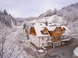 Uma casa de madeira de dois andares com telhado inclinado, rodeada por árvores e colinas cobertas de neve.