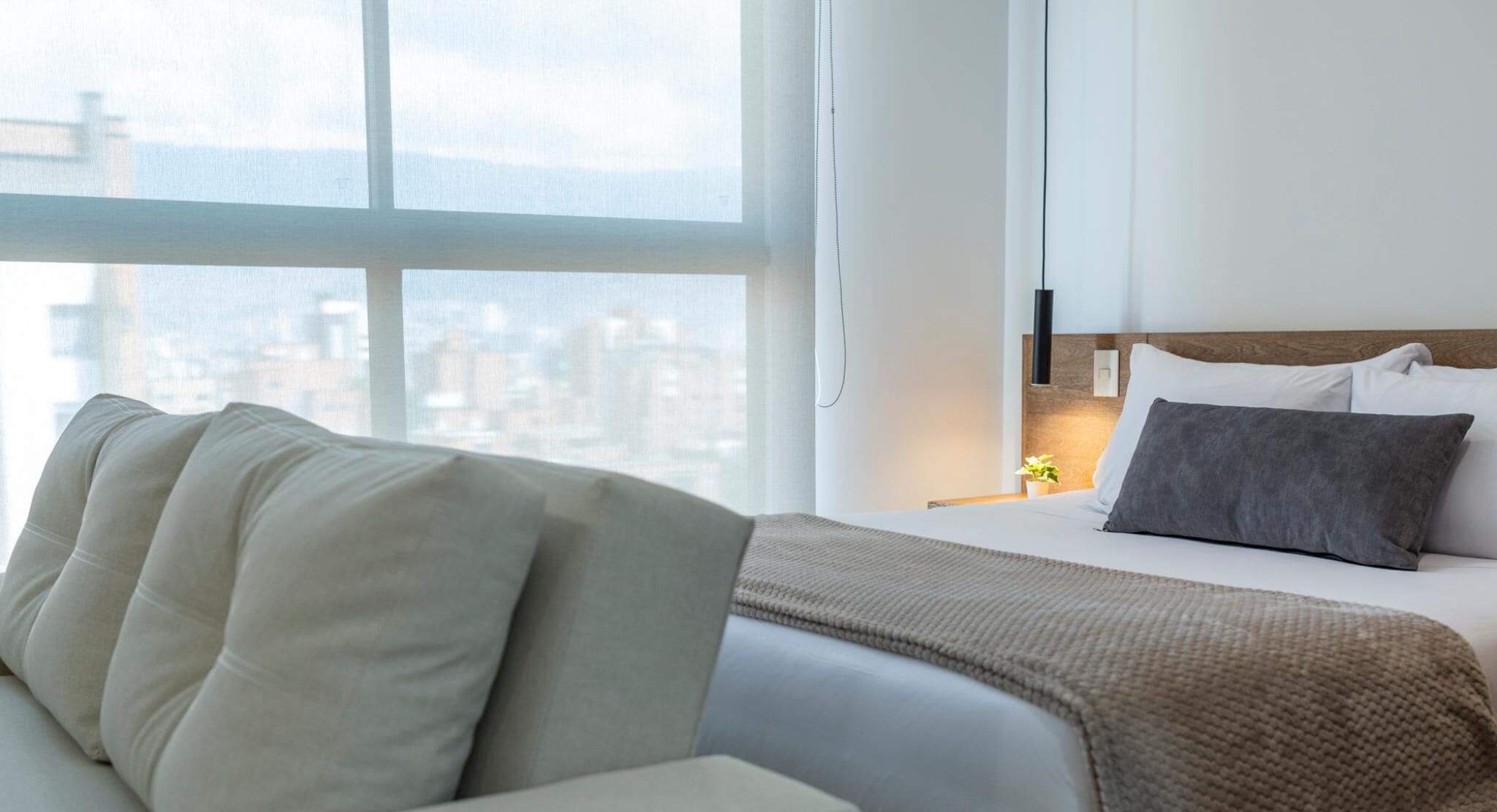 Quarto de hotel moderno com uma janela grande, uma cama com roupa de cama branca e cinza, e um sofá com almofadas em tons claros.