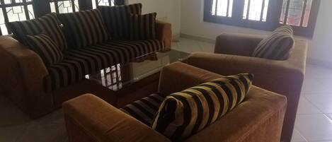 Uma sala de estar com sofá de listras, mesa de centro de vidro e poltrona marrom.