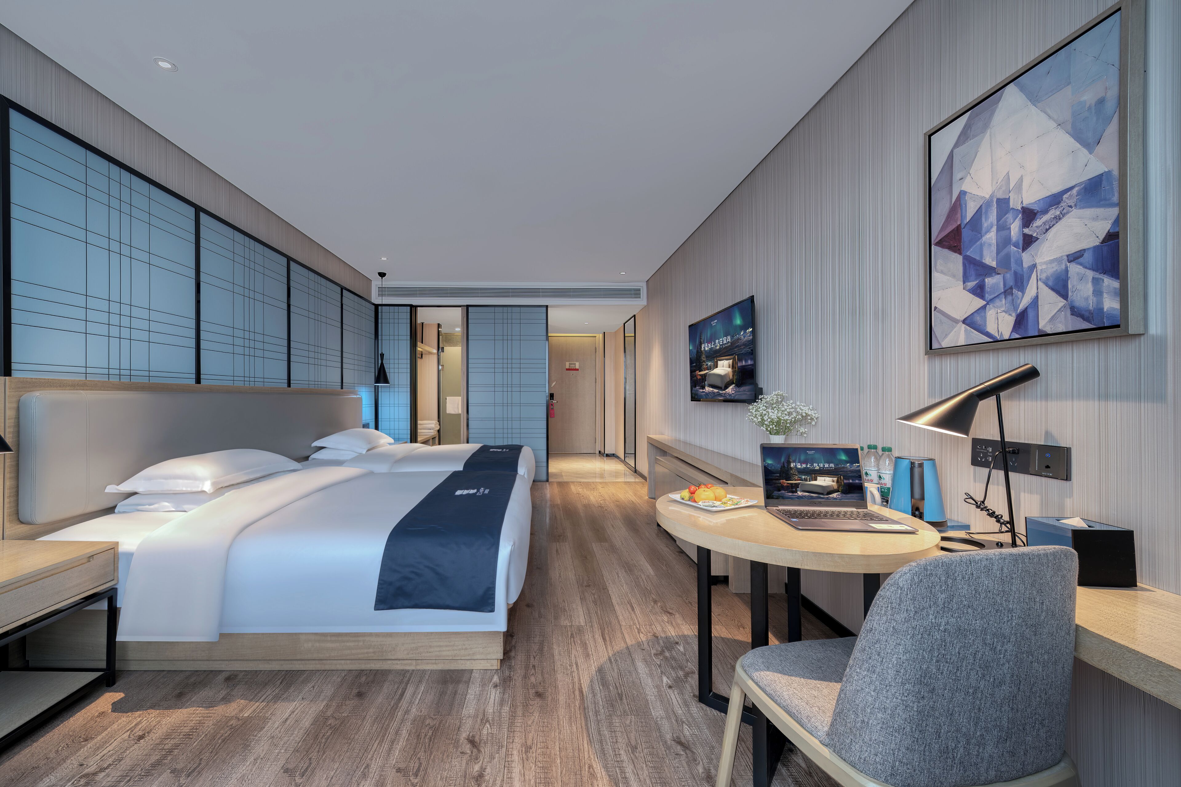 Quarto de hotel moderno com cama, escrivaninha e cadeira. Há uma janela ampla e um quadro na parede.