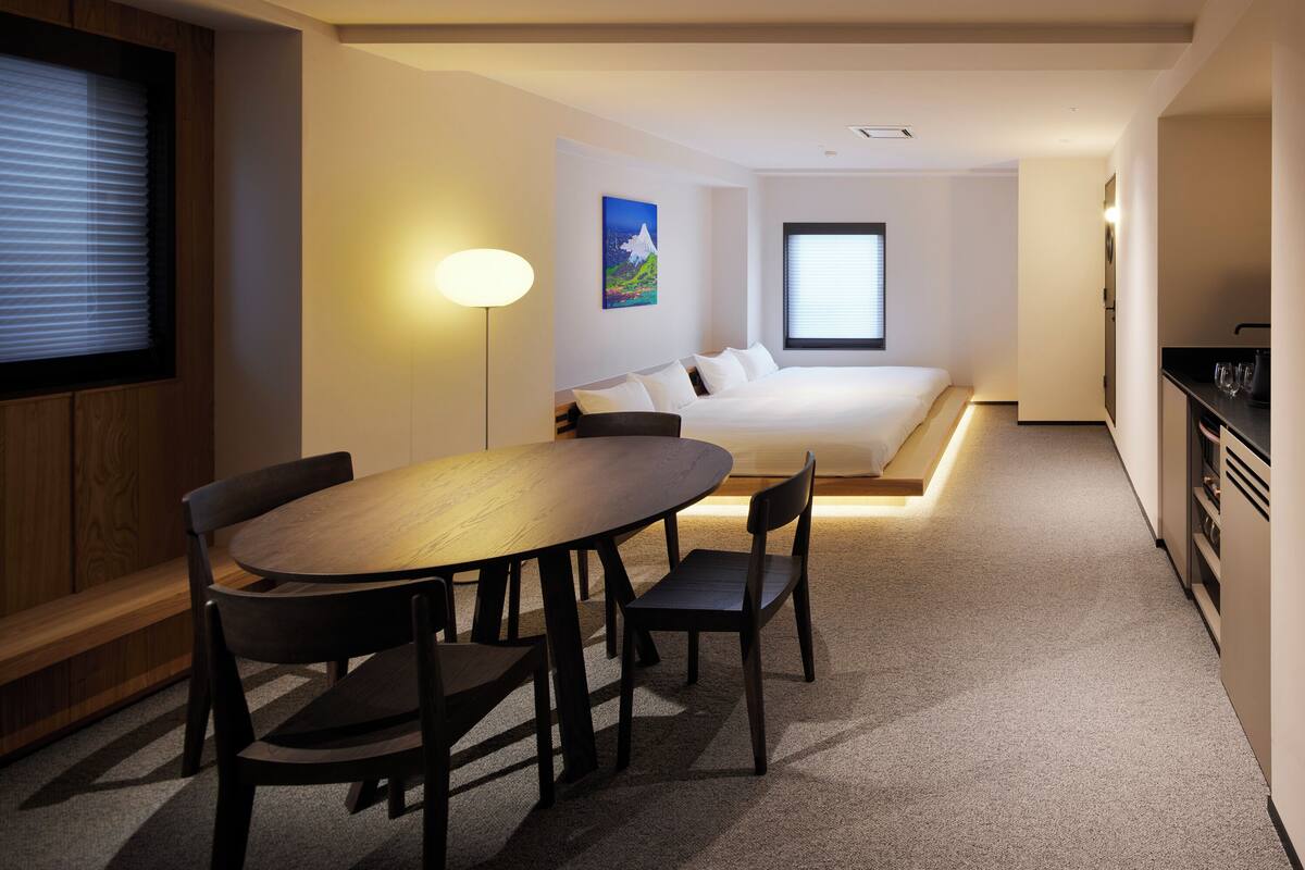 Quarto de hotel compacto com cama, área de refeições com mesa e cadeiras, pequena cozinha e janela com persianas.