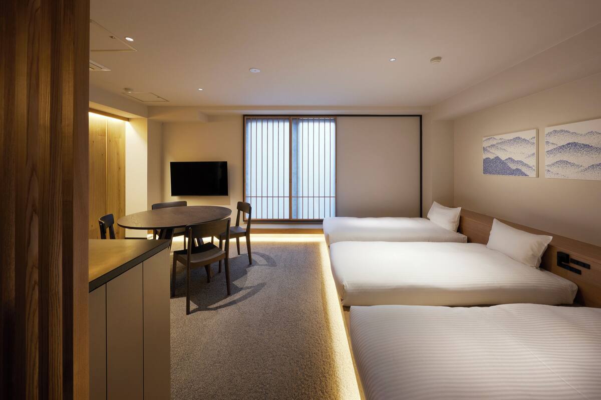 Quarto de hotel com duas camas, área de refeições com mesa e cadeiras, televisão e obras de arte na parede.