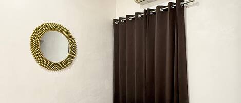 Uma casa de banho com duas cadeiras, uma mesa pequena com uma toalha e uma garrafa, um espelho, uma cortina e um aparelho de ar condicionado.