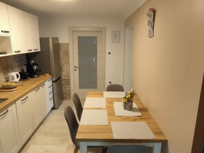 Cozinha compacta com armários brancos, cafeteira e torradeira. Há uma mesa de jantar com cadeiras e um vaso de flores.