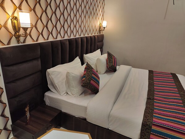 Quarto de hotel com cabeceira escura, roupa de cama branca e manta listrada colorida. Há duas luminárias fixadas na parede e uma mesa pequena com um objeto decorativo.
