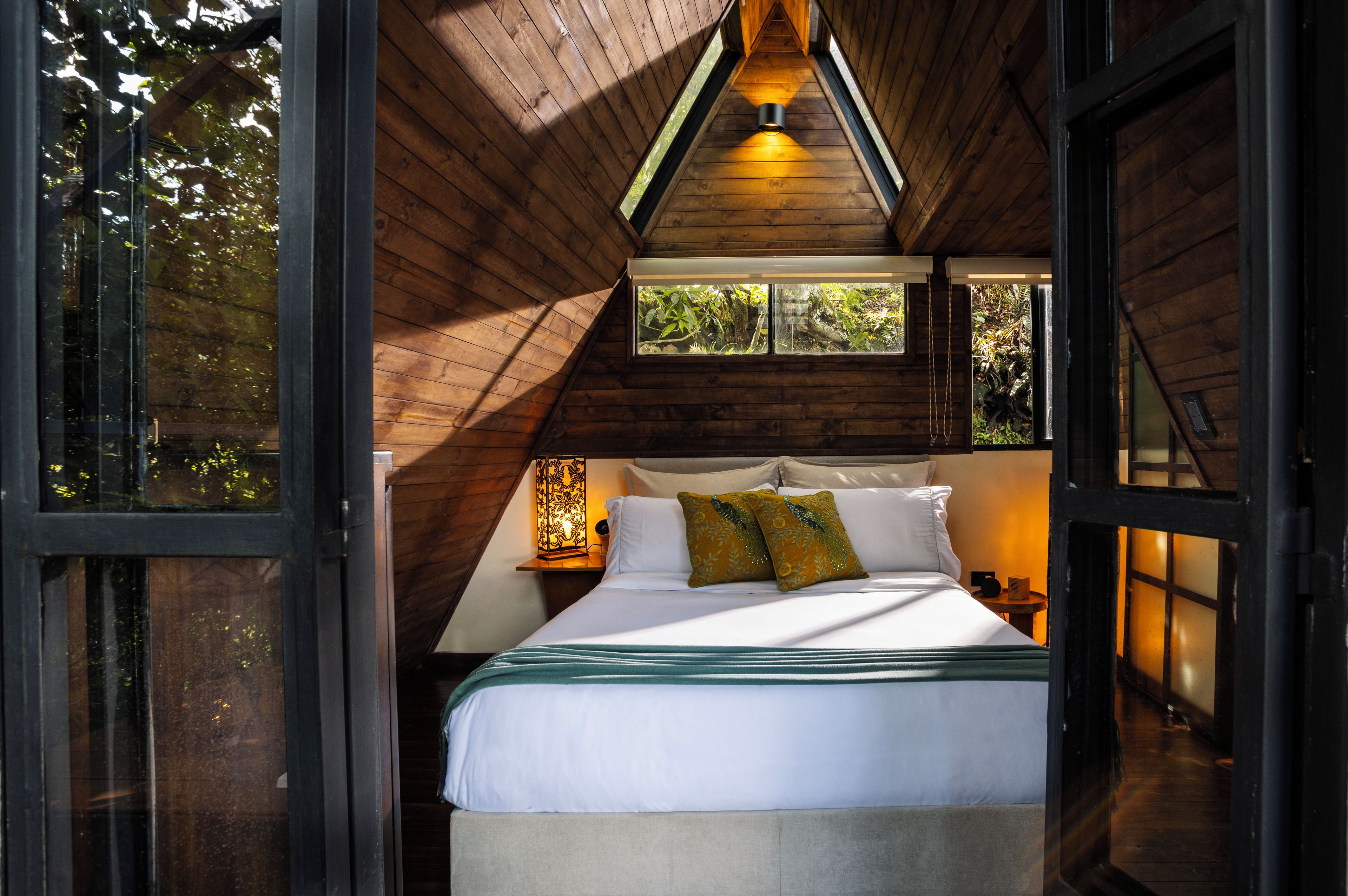 Quarto com teto de madeira, cama com roupa de cama branca e almofadas decorativas, mesa de cabeceira com abajur e janela com vista para o verde.