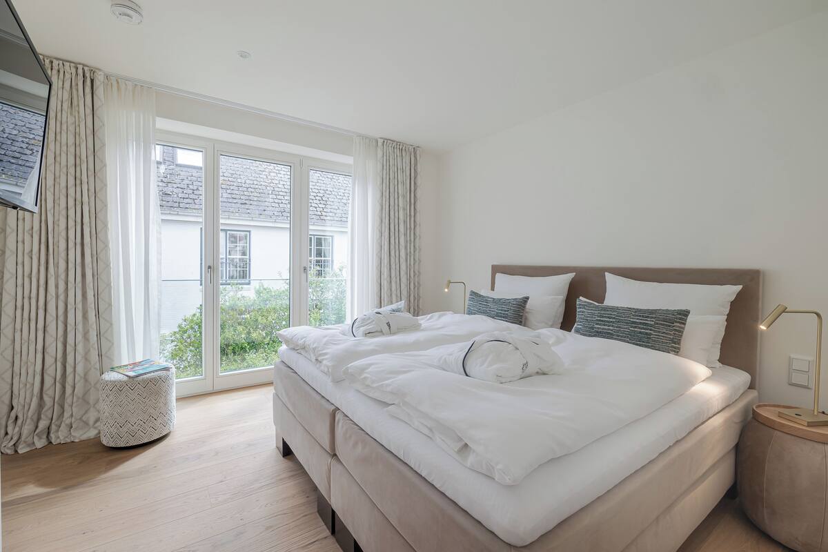 Quarto de hotel com uma cama grande, uma mesa de cabeceira com abajur, uma janela com cortinas e vista para o exterior.