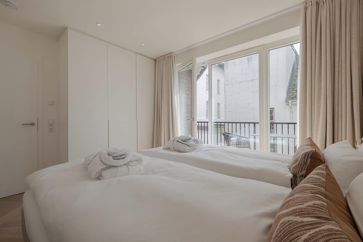 Quarto de hotel com duas camas, uma janela grande com cortinas, um guarda-roupa branco e vista para um edifício.