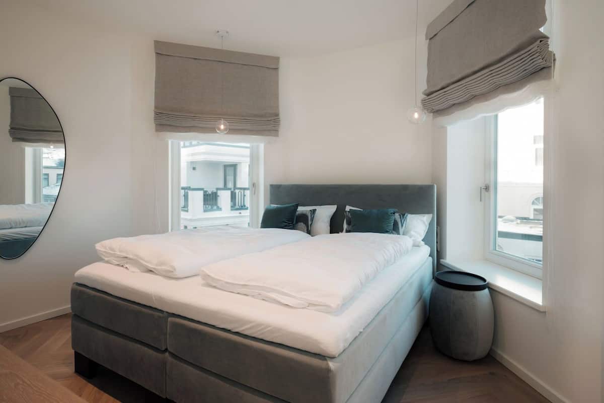 Quarto moderno com cama grande, espelho redondo, vista para a varanda e janela com persianas.