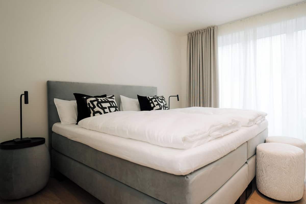 Quarto moderno com cama cinza, roupa de cama branca e luminária de cabeceira.